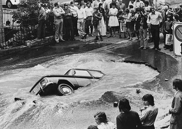 A crowd of spectators gather around to watch a car sinking into a large ...